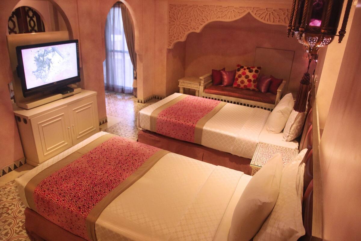 Quarto de hotel com duas camas, televisão e um lustre decorativo.