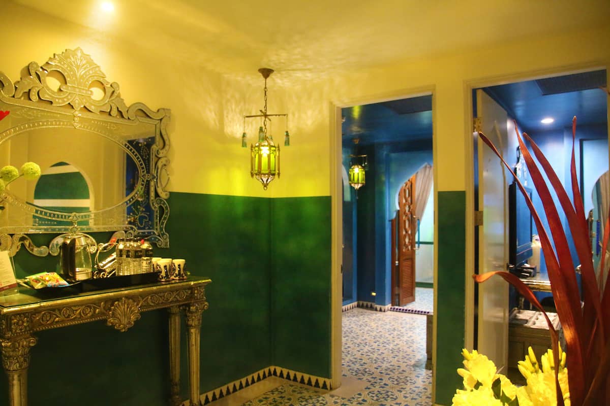 Quarto com parede verde e amarela, um espelho grande e decorado, uma mesa com bandeja e um candeeiro suspenso.