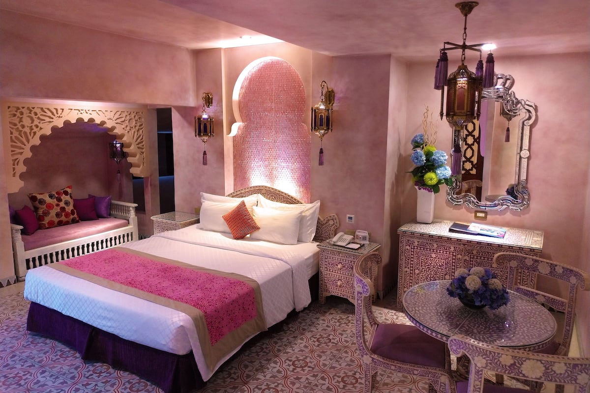 Quarto em estilo tradicional marroquino, com cama, sofá, painéis decorativos na parede, um candelabro e uma pequena mesa redonda com cadeiras.