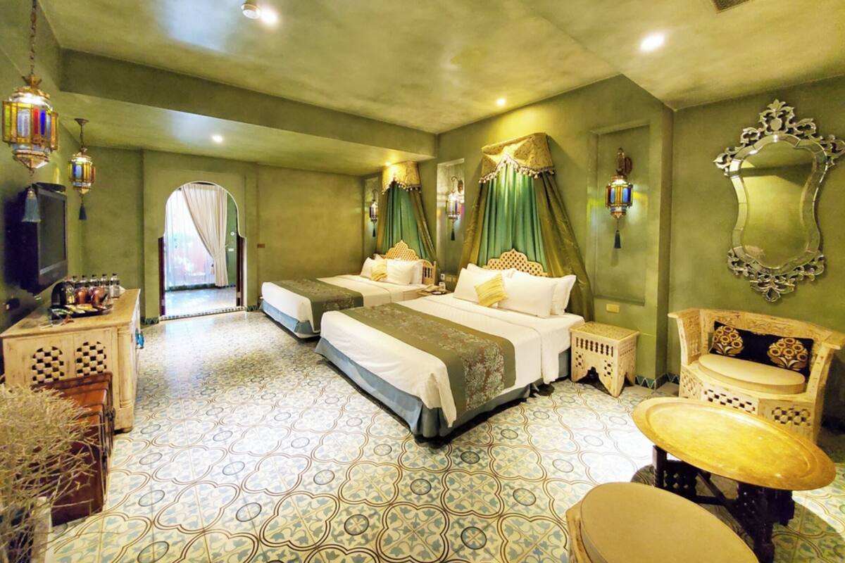 Quarto de hotel com duas camas, um sofá, uma mesa pequena e um espelho decorativo.