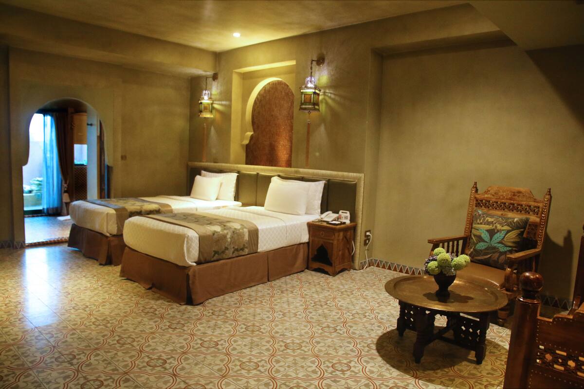 Quarto de hotel com uma cama grande, duas mesas de cabeceira, uma mesa de centro redonda e uma cadeira com almofada decorativa.