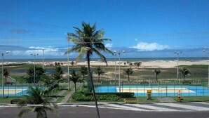 Vista da praia com quadra esportiva cercada, palmeiras e céu claro.