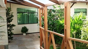 Uma área de varanda coberta com estrutura de pérgola de madeira, plantas em vasos e vista para o verde ao redor.