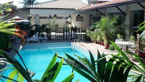 Piscina com espreguiçadeiras e mesas, rodeada por plantas em vasos e um edifício com telhado em azulejos.