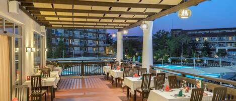 Uma área de refeições ao ar livre, com mesas e cadeiras, piscina ao fundo e vista para edifícios residenciais.