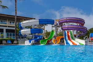 Um parque aquático com vários toboáguas em uma área de piscina, cercado por um edifício com varandas e uma palmeira.