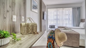 Quarto de hotel moderno com parede de destaque em madeira, uma janela grande com cortinas finas, cama com cobertura acolchoada, TV fixada na parede e uma mesinha com uma bolsa tecida e óculos de sol.