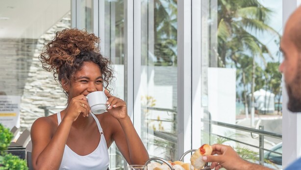 Uma área de refeições moderna com mesas e cadeiras de vidro, vista para palmeiras do lado de fora e um ambiente descontraído com pratos de comida e bebidas.
