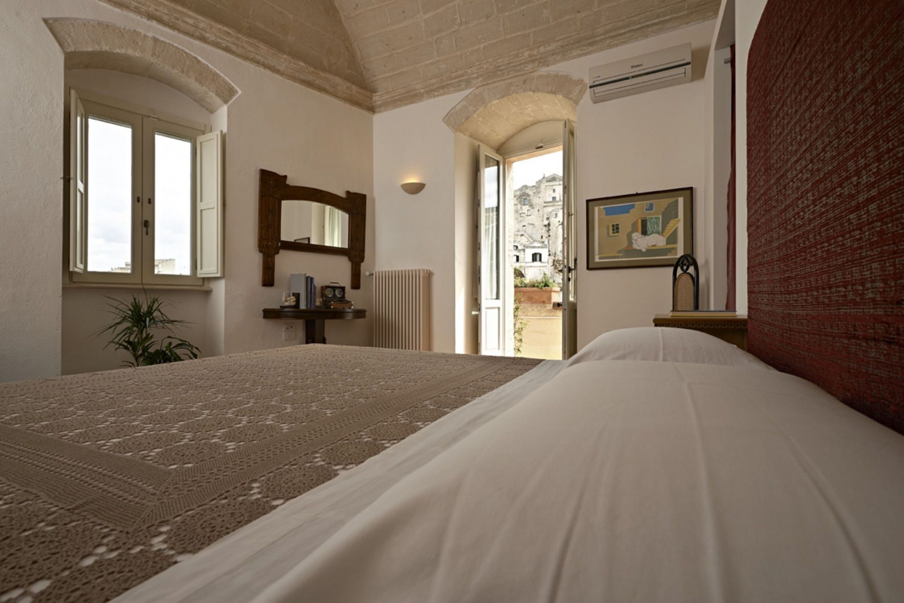 Quarto com uma cama grande, uma mesinha com luminária, um espelho e vista para edifícios por uma porta aberta.