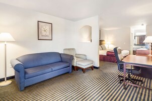 Quarto de hotel com sofá azul, uma poltrona pequena, uma mesa de jantar com uma cadeira e um tapete estampado.