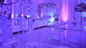 Salão de banquetes com palco, arranjos florais brancos e cadeiras transparentes.