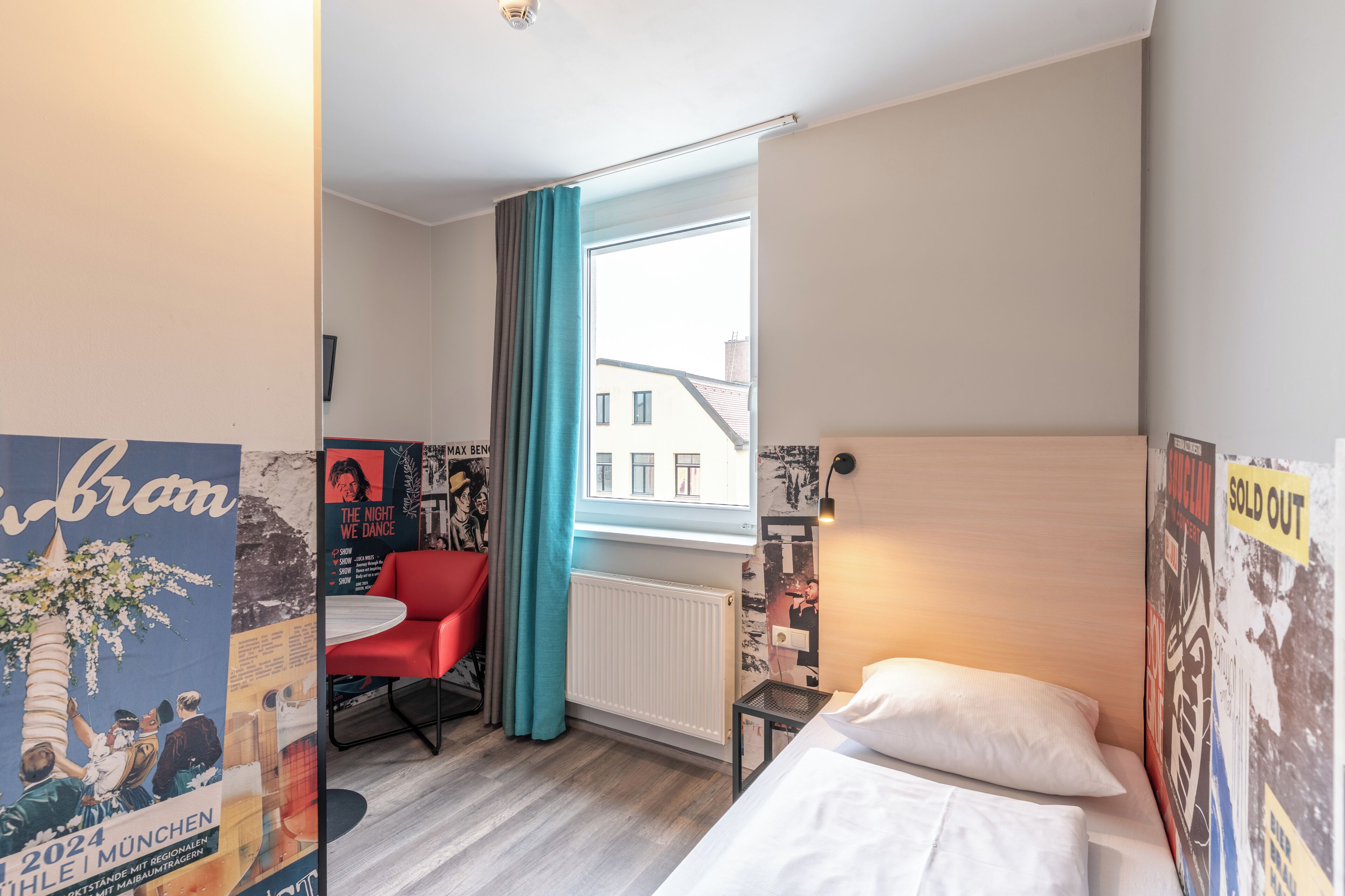 Quarto de hotel com cama, uma cadeira vermelha, janela com cortinas e um cartaz na parede.