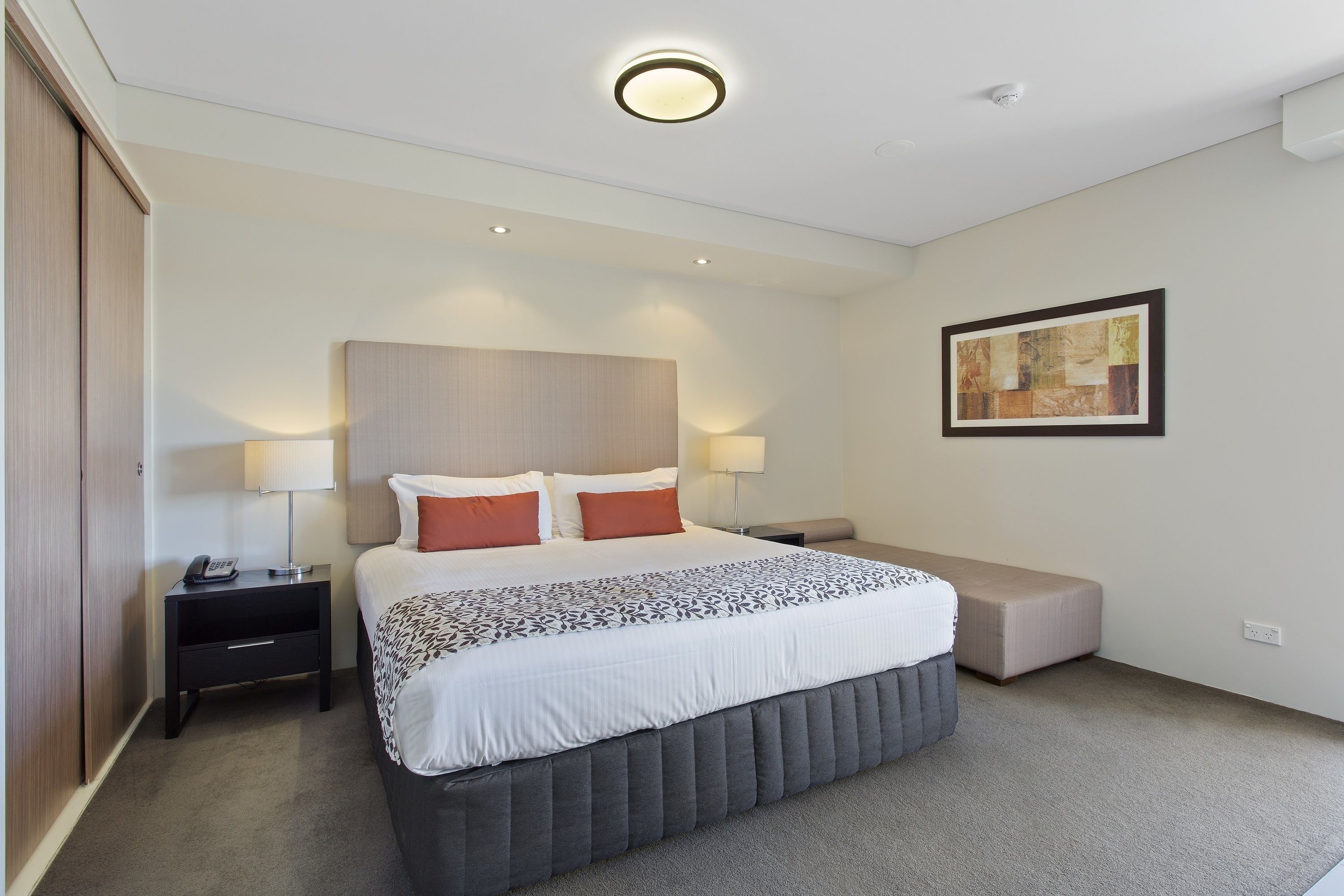 Quarto de hotel com cama grande, luminárias de cabeceira, mesa de cabeceira com telefone, um banco e uma obra de arte emoldurada na parede.