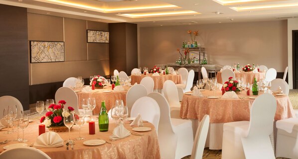 Salão de banquetes com mesas redondas dispostas para um evento formal, com cadeiras brancas, louças e centros de mesa florais.