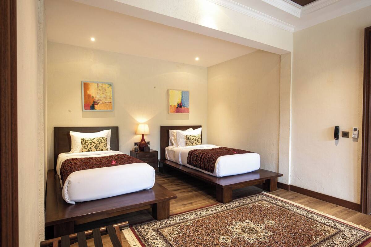 Quarto de hotel com duas camas, um banco de madeira, um tapete estampado e dois quadros na parede.