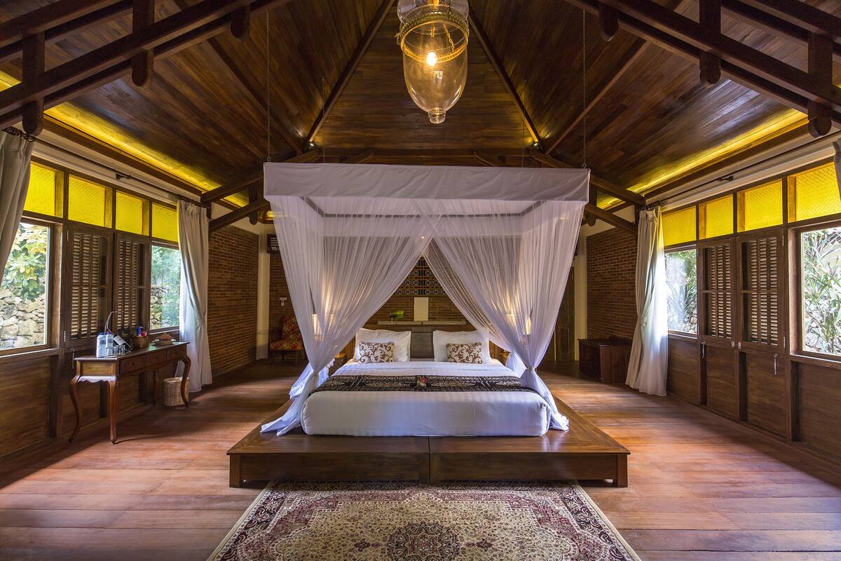 Um quarto espaçoso com cama de dossel, piso de madeira e grandes janelas com persianas.