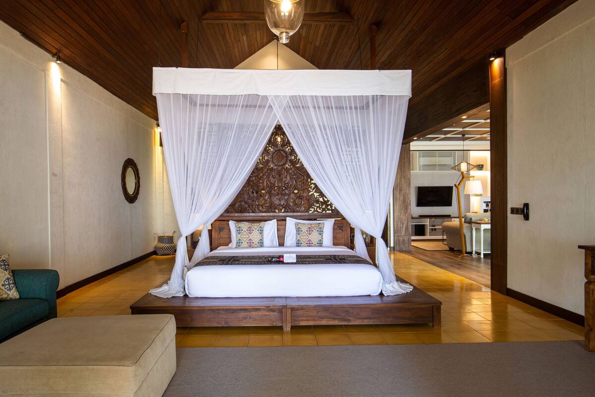 Quarto com cama de dossel, sofá, piso de madeira e televisão ao fundo.
