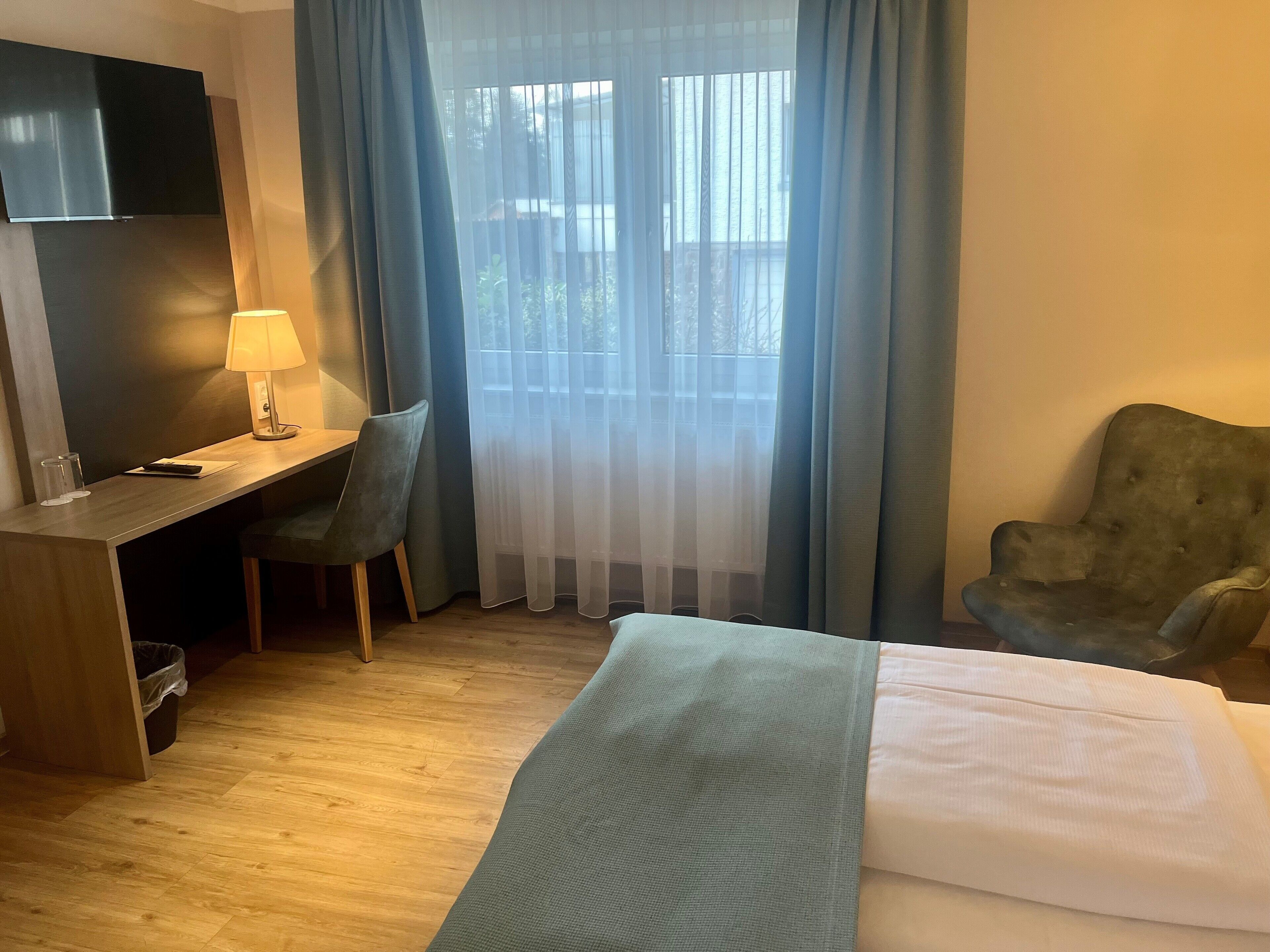 Quarto de hotel com uma cama, uma mesa com abajur, duas cadeiras e uma televisão.