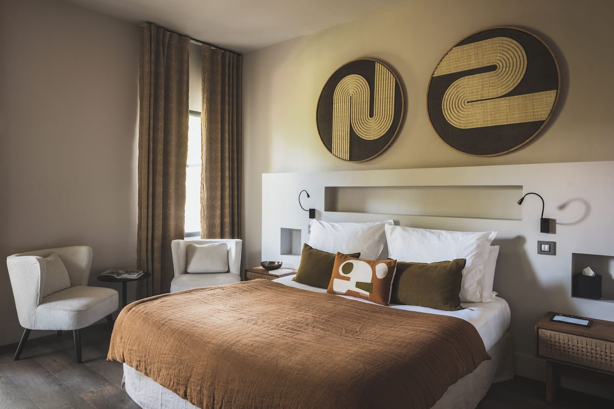 Quarto de hotel com uma cama grande, duas cadeiras laterais, uma mesa de cabeceira e uma obra de arte pendurada na parede.
