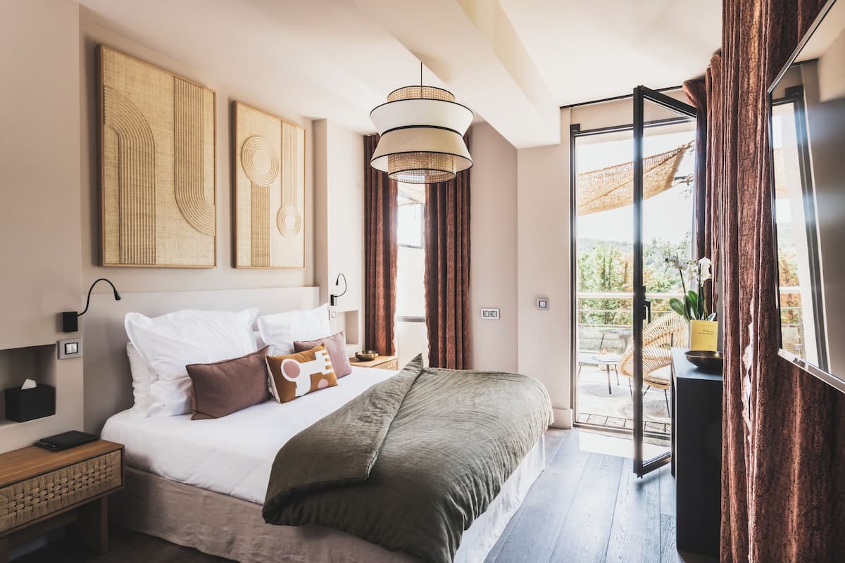 Quarto com cama, mesa de cabeceira, abajur, obra de arte e vista através de uma porta de vidro.