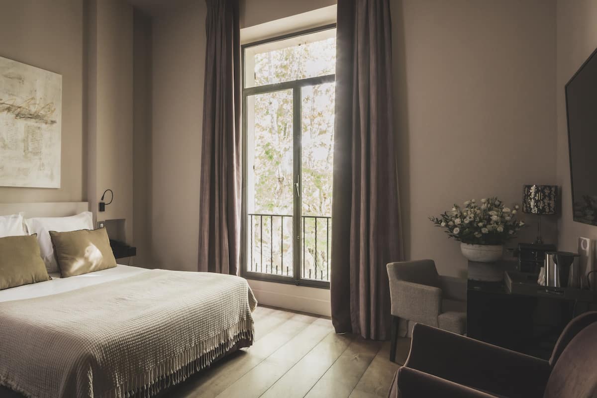 Quarto com uma cama grande, janela com cortinas, mesa de cabeceira, cadeira e um quadro na parede.