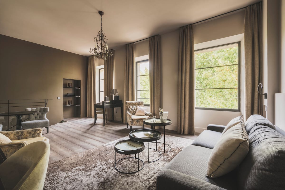 Uma sala de estar com sofá, poltronas, mesa de centro, lustre e grandes janelas com cortinas.