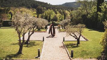 Uma trilha de cascalho serpenteia por um jardim com árvores bem aparadas e uma paisagem montanhosa ao fundo.