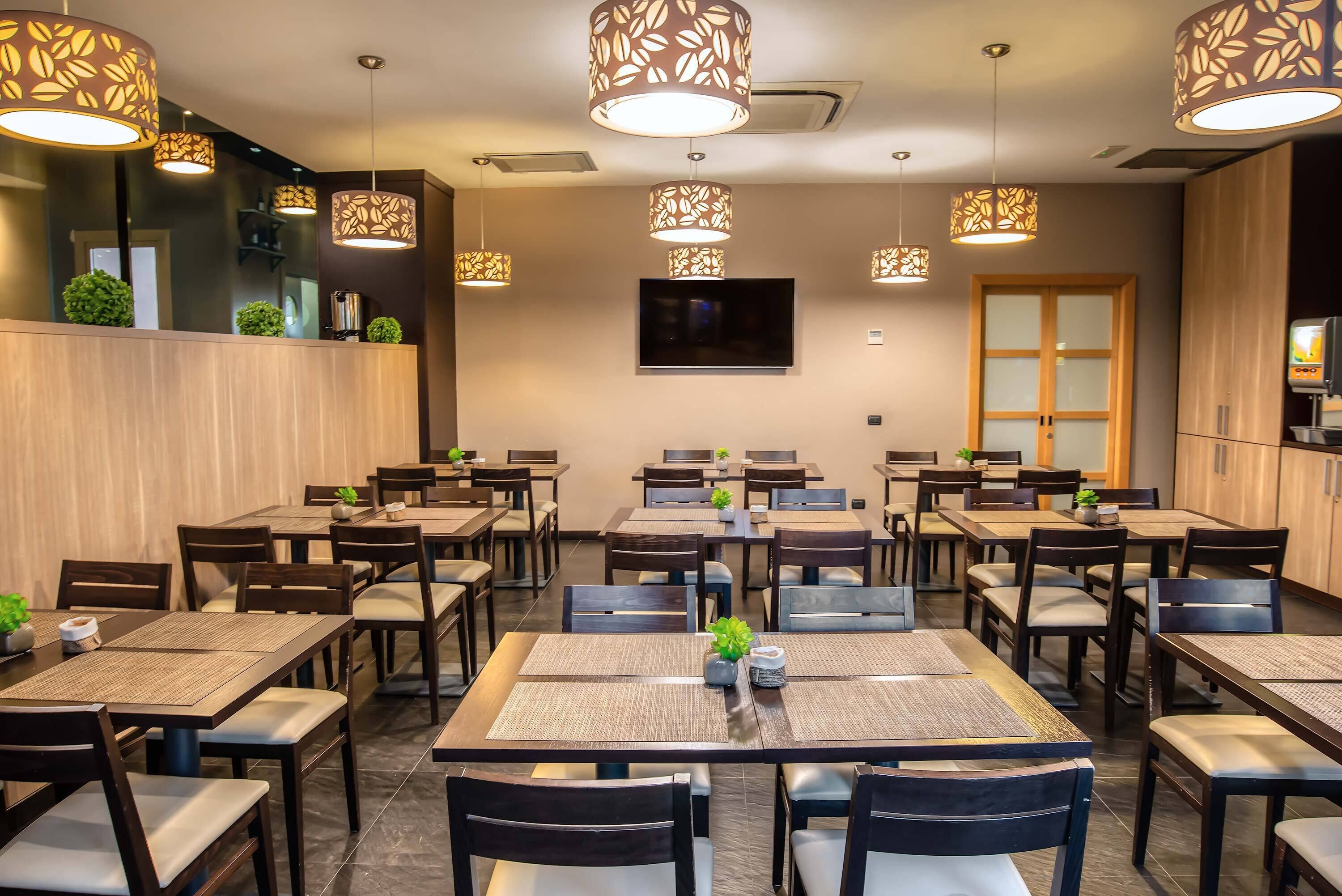 Um restaurante com várias mesas e cadeiras, luminárias decorativas penduradas, uma televisão de tela plana na parede e um balcão de madeira com plantas.