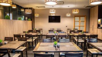 Um restaurante com várias mesas e cadeiras, luminárias decorativas penduradas, uma televisão de tela plana na parede e um balcão de madeira com plantas.