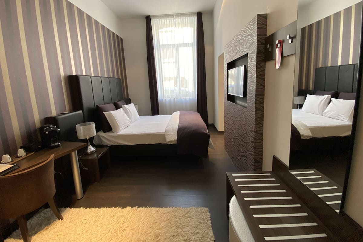 Quarto de hotel com cama, mesas de cabeceira, escrivaninha, espelho e janela com cortinas.