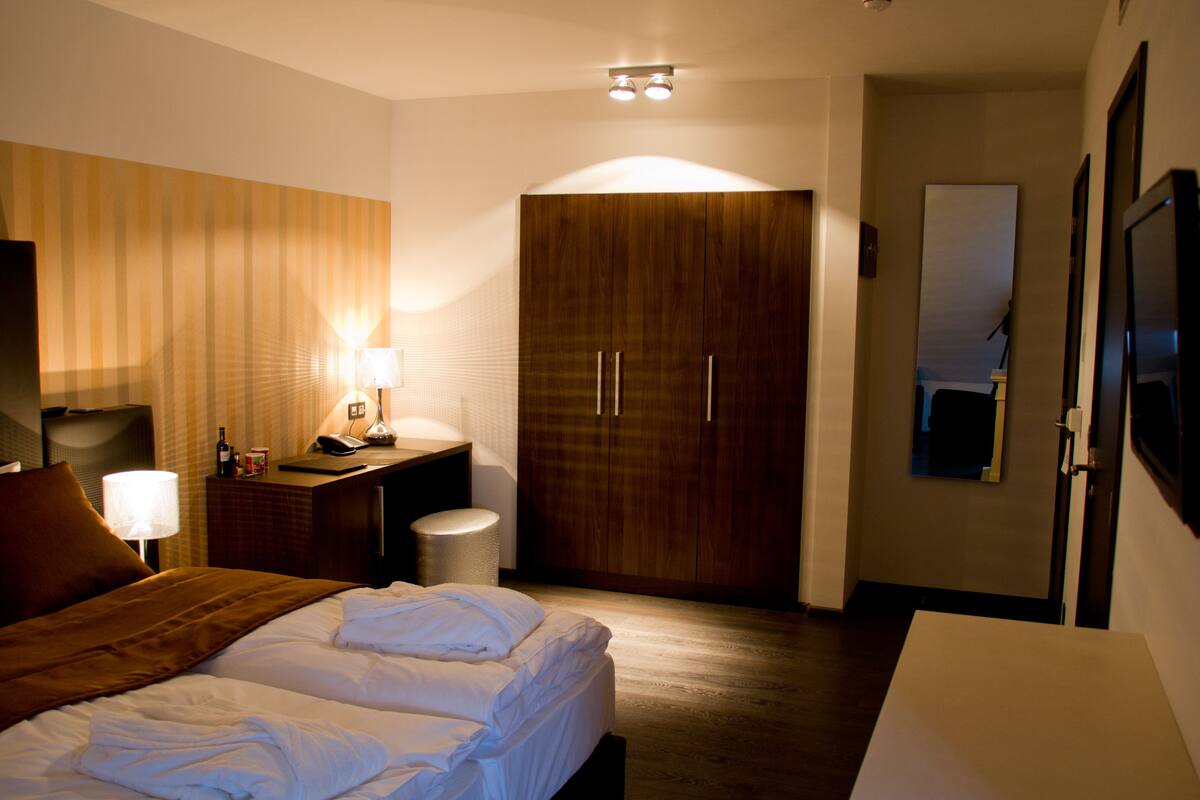 Quarto de hotel com cama, mesa de cabeceira, luminária, guarda-roupa e espelho.
