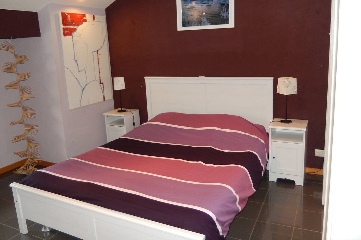 Quarto com cama branca, roupa de cama listrada em roxo e rosa, duas mesas de cabeceira com luminárias, um quadro na parede e teto com vigas de madeira.