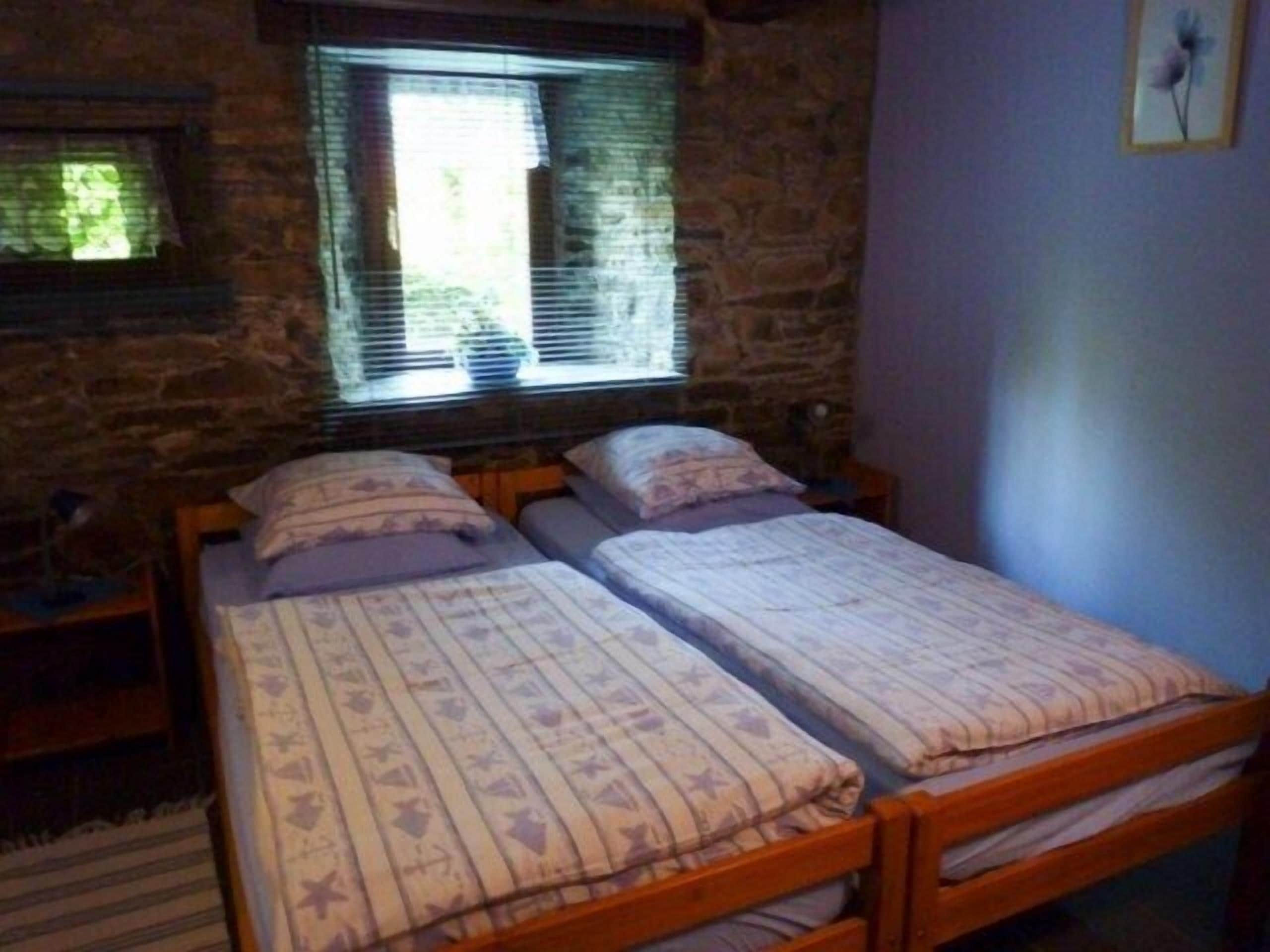 Quarto com duas camas de solteiro, parede de pedra, janela com persianas e um quadro pendurado na parede.