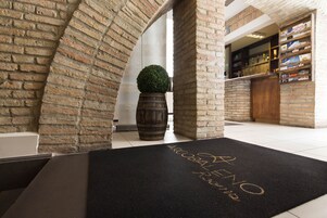 Uma área de recepção com um tapete preto, paredes de tijolo, um barril de madeira e uma pequena planta em vaso.