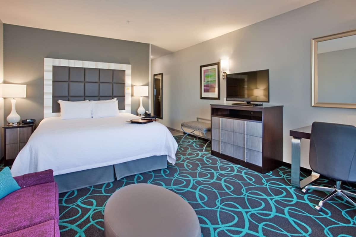 Quarto de hotel com uma cama grande, uma escrivaninha com computador, uma televisão e uma cadeira.