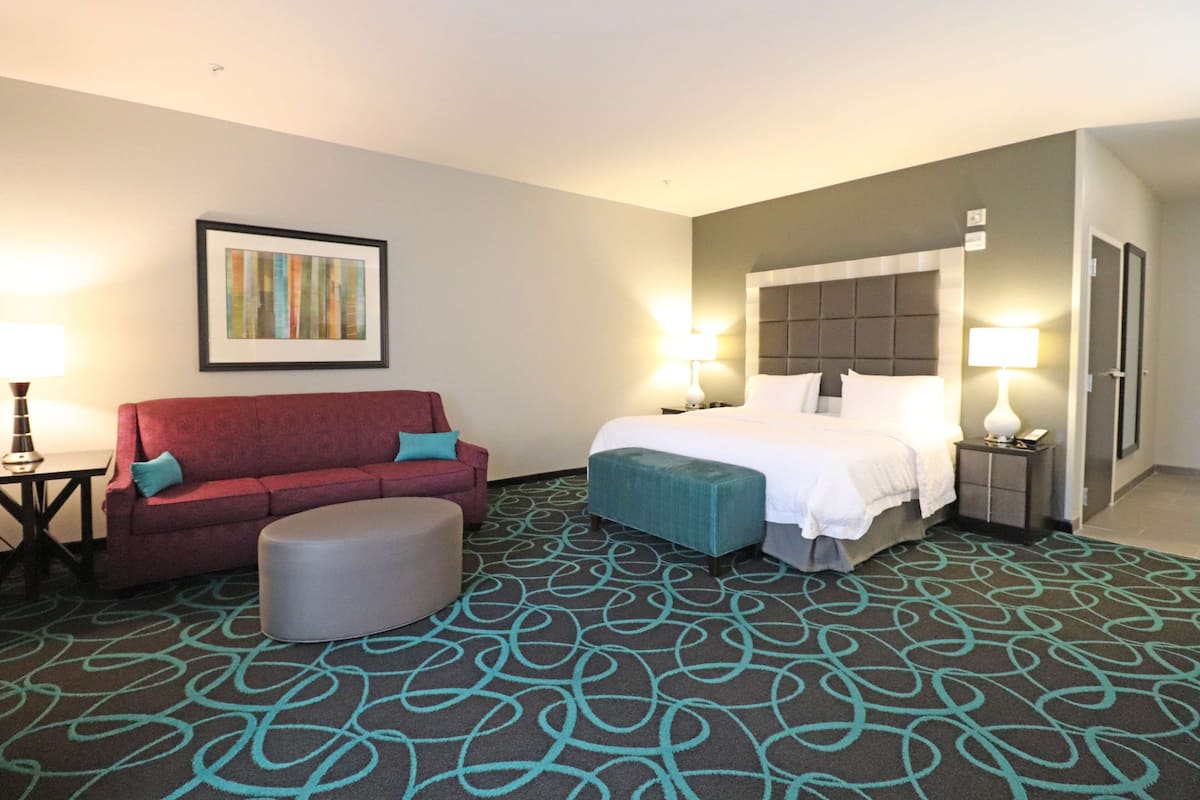 Quarto de hotel com uma cama, um sofá vermelho, um banquinho cinzento, uma obra de arte emoldurada na parede e um tapete estampado.