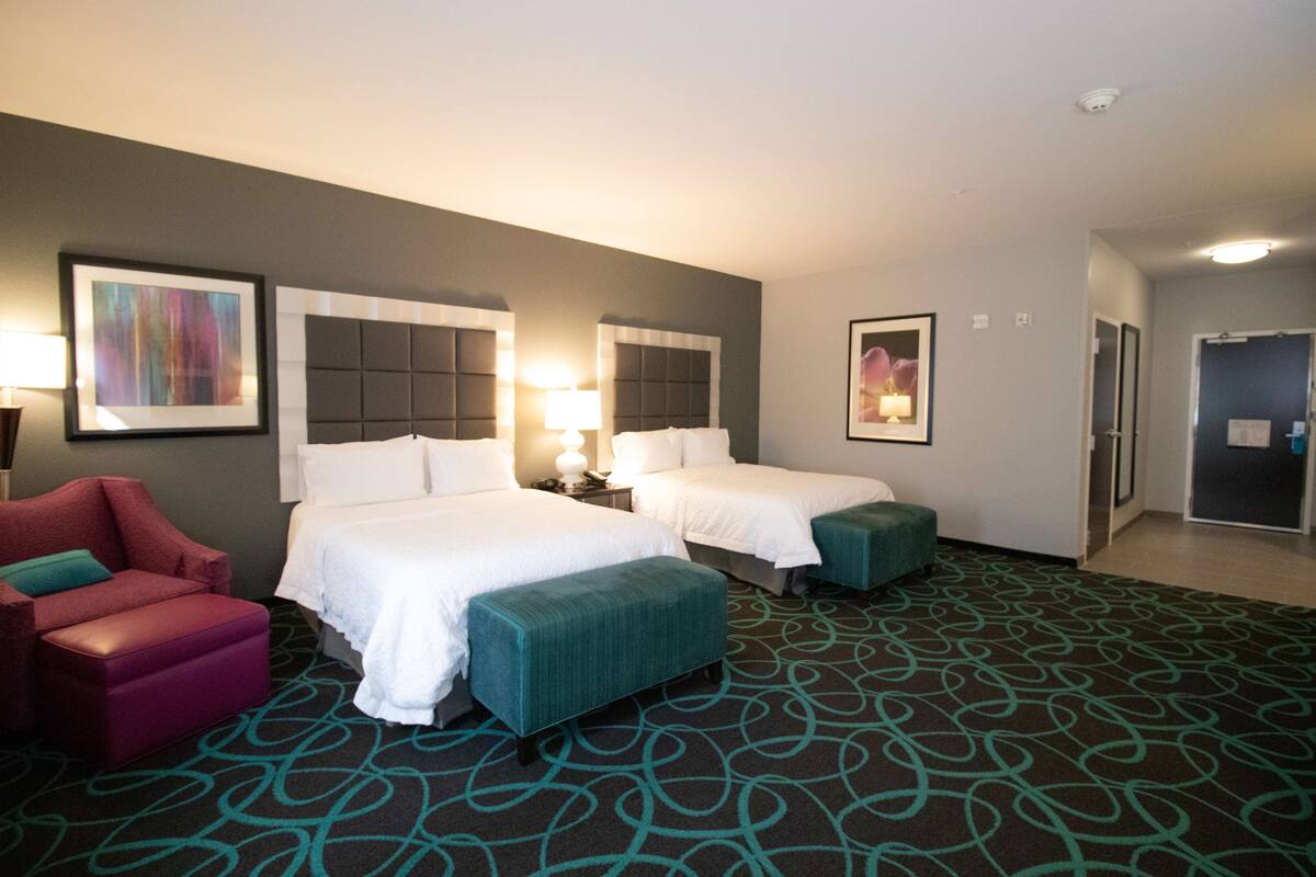 Quarto de hotel com duas camas, uma poltrona vermelha e um banquinho azul-esverdeado.