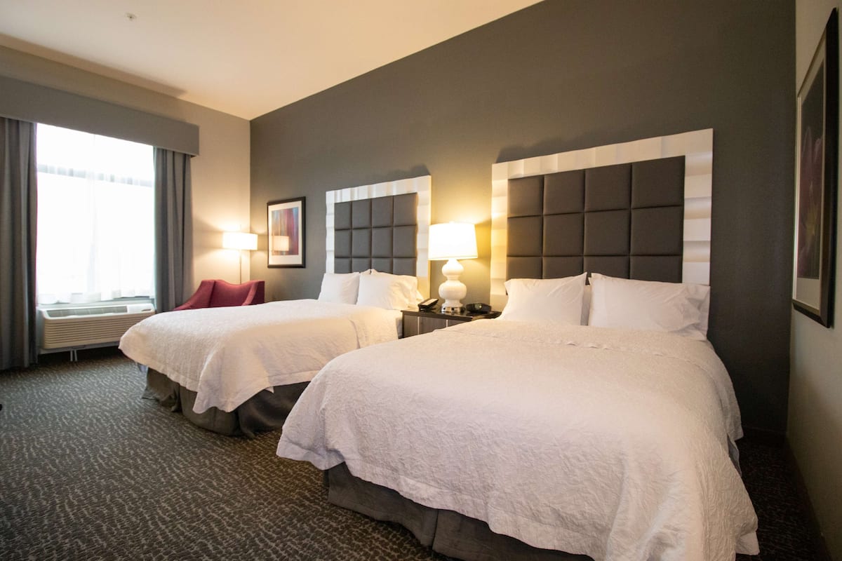 Quarto de hotel com duas camas, uma janela grande com cortinas e chão coberto de carpete.