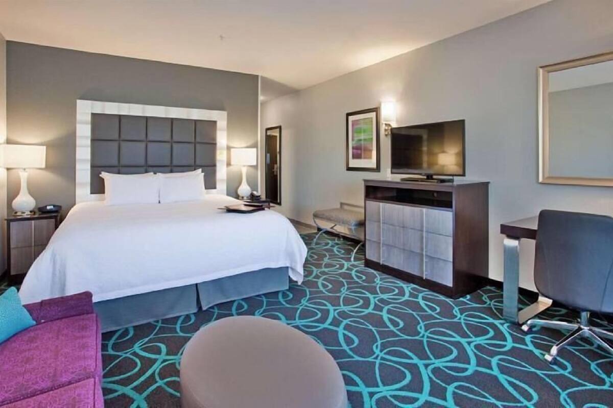 Quarto de hotel com cama, escrivaninha, cadeira, televisão e espelho.