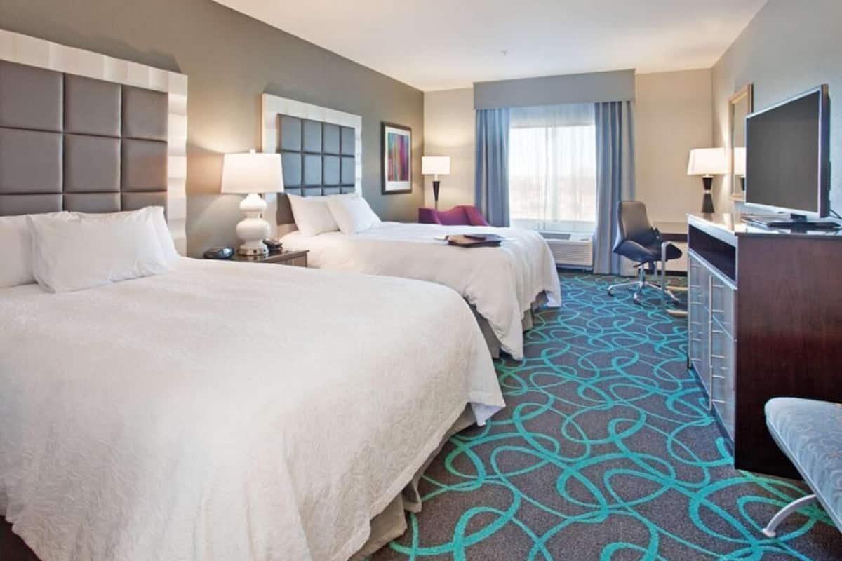 Quarto de hotel com duas camas, uma escrivaninha, televisão e janela com cortinas.