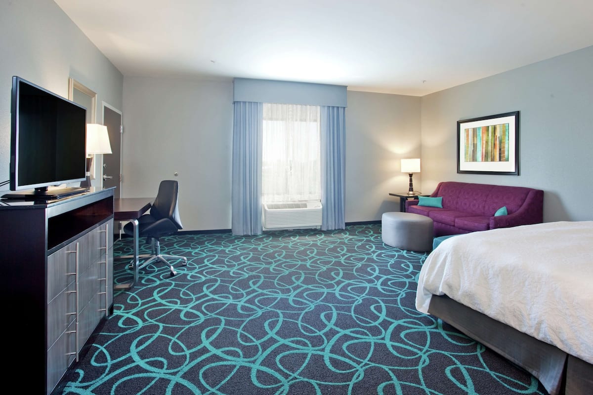 Quarto de hotel com cama, escrivaninha com televisão, cadeira, sofá, abajur e janela com cortinas.