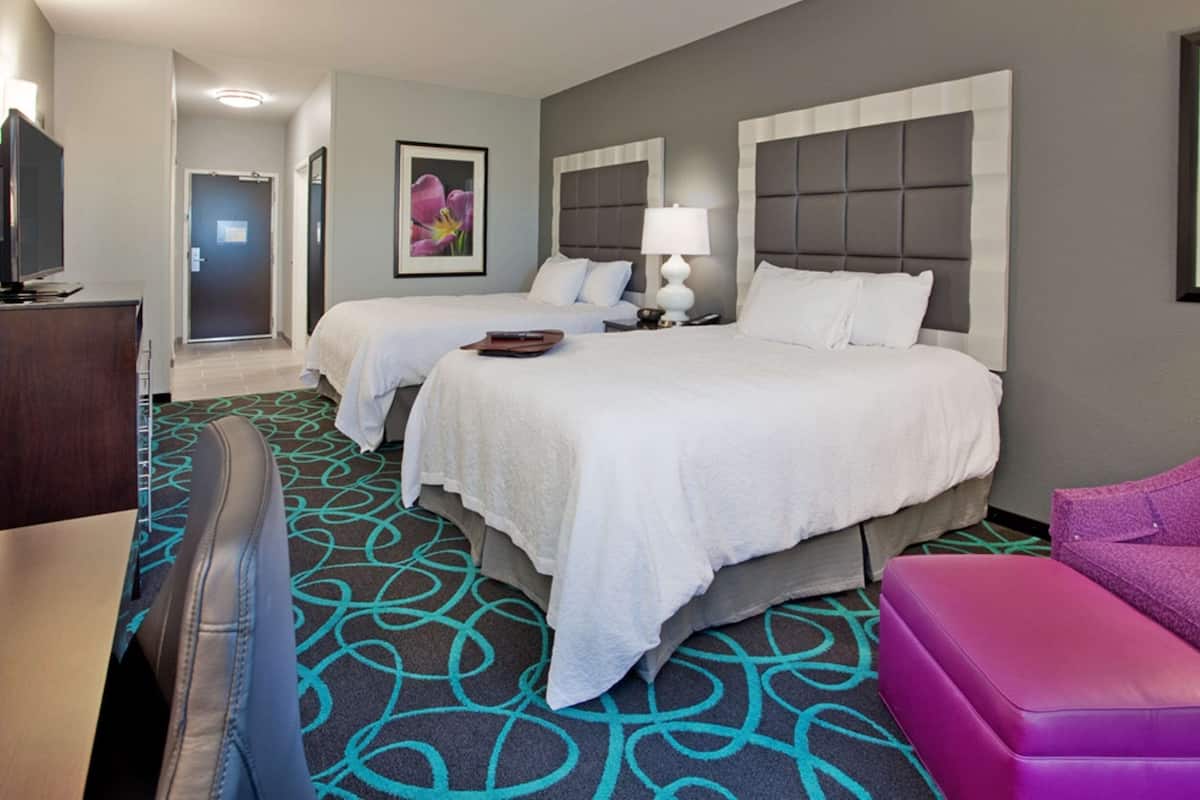 Quarto de hotel com duas camas, televisão, escrivaninha e um tapete colorido.