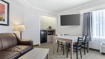 Quarto de hotel com sofá, mesa de jantar com cadeiras, TV de tela plana na parede, área de cozinha com geladeira e micro-ondas, e janela com cortinas.