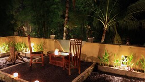 Uma área externa com assentos, incluindo duas cadeiras de madeira e uma mesa, cercada por plantas em vasos e palmeiras.
