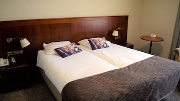 Quarto de hotel com uma cama de casal, duas almofadas, mesa de cabeceira com abajur, mesa redonda pequena e telefone.