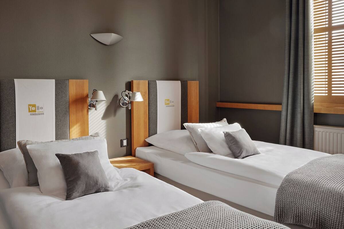 Quarto de hotel com duas camas, cabeceira de madeira, luminária na parede e janela com persianas.
