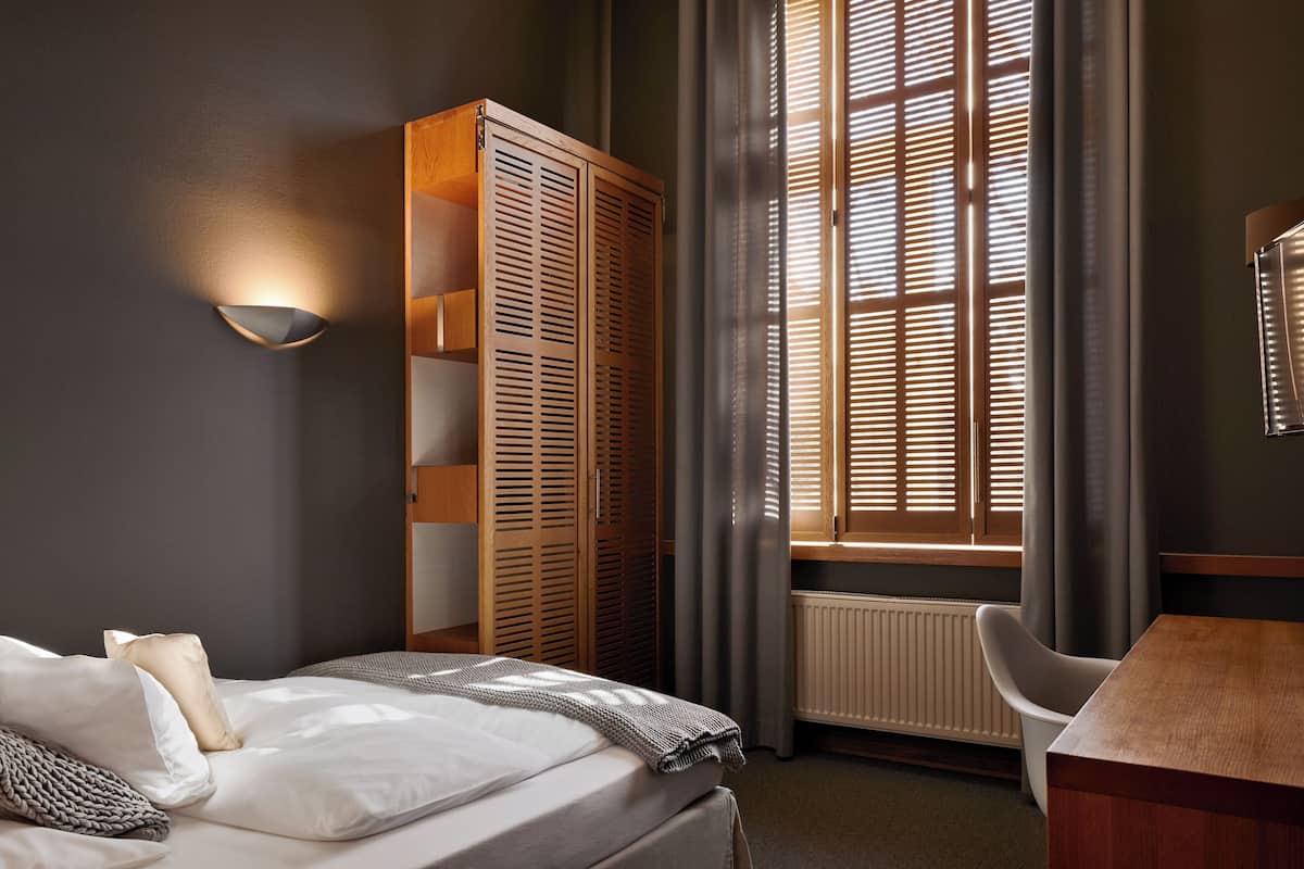Quarto com cama, guarda-roupa de madeira, escrivaninha e janela com persianas.