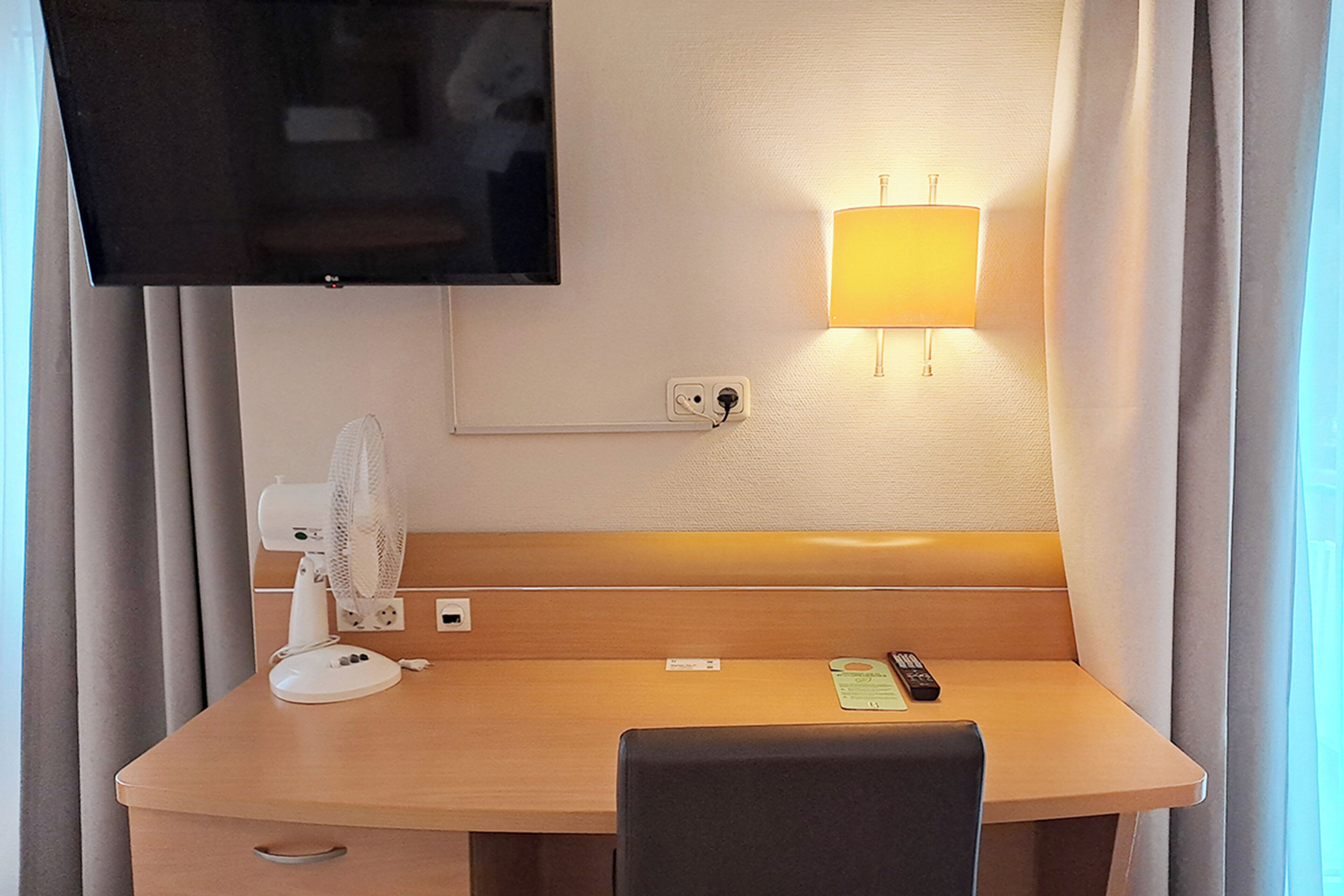 Quarto de hotel com escrivaninha de madeira, televisão na parede, luminária de cabeceira e ventilador.
