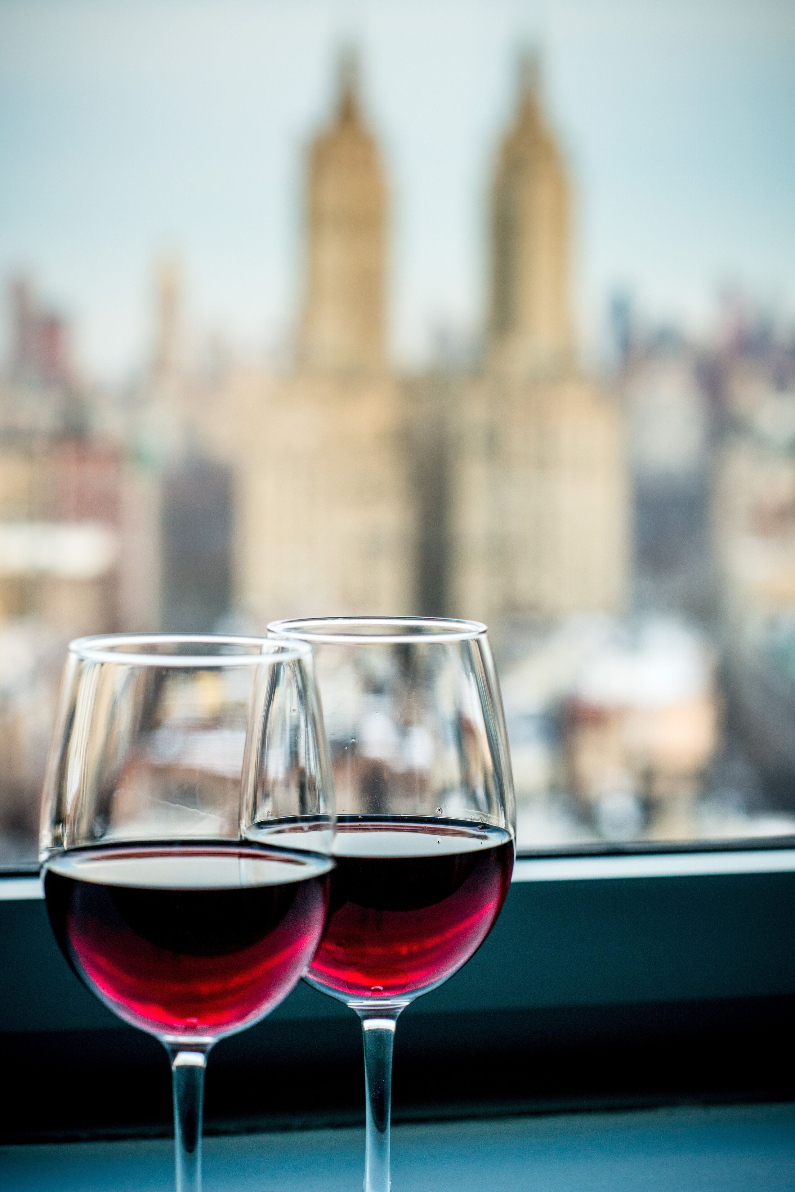 Duas taças de vinho com líquido vermelho, posicionadas em frente a uma janela com vista para um horizonte urbano e edifícios imponentes.