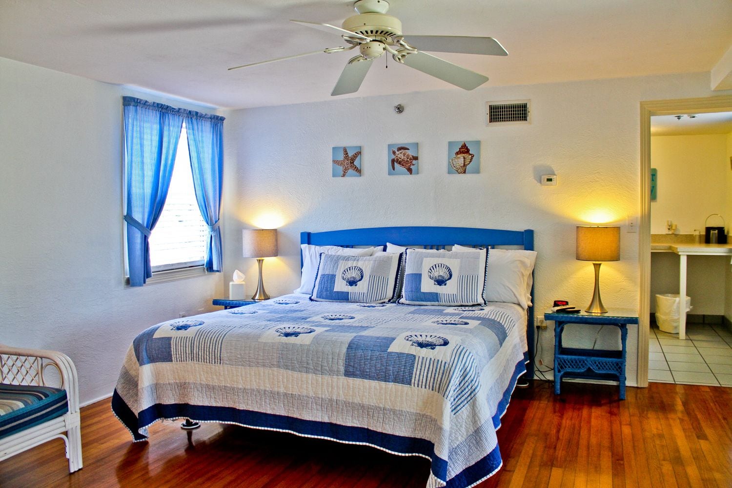 Quarto com cama, duas mesas de cabeceira, ventilador de teto, janela com cortinas e obras de arte nas paredes.